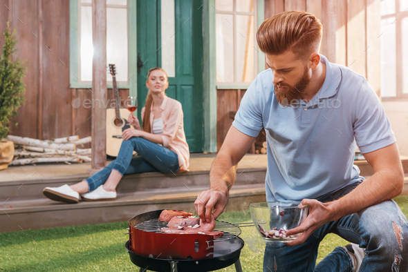 bearded man roasting meat on barbecue grill with woman with wine behind ...