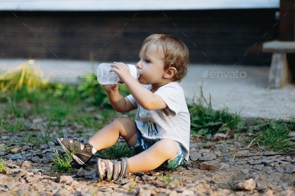 Cute little boy sipping water from baby bottle sitting on the ground ...