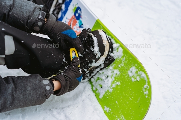 little boy sitting on snow putting his feet in snowboard bindings ...