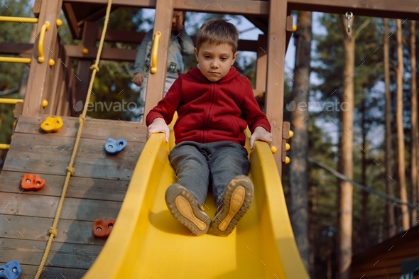 Cute little boy sliding down plastic slide on playground in countryside ...