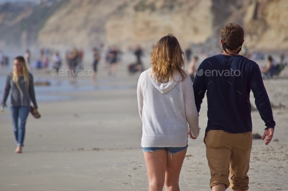 Happy cute couple walking along the beach holding hands with beachgoers ...