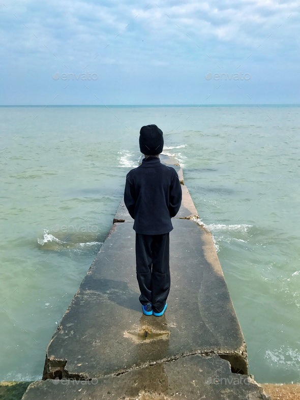 Young boy facing away all dressed in black on a pier looking at Lake ...