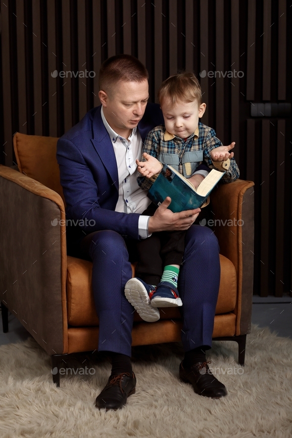 son and dad reading a book sitting in a chair Stock Photo by katyatsiganok