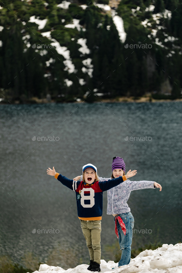 children are standing on the melted snow, overlooking a mountain lake ...