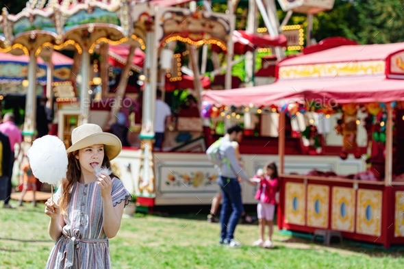 A girl in a hat and a dress eating cotton candy on a hot summer day at ...