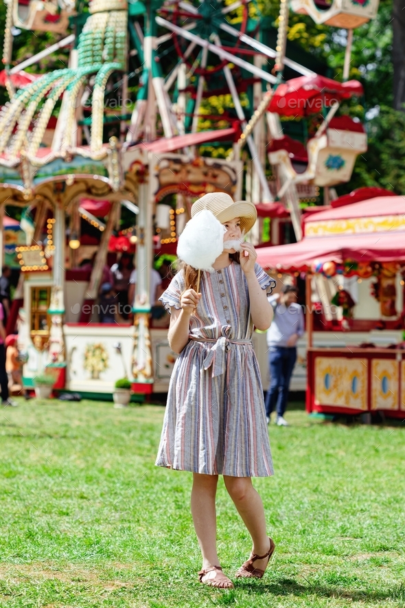 A girl in a hat and a dress eating cotton candy on a hot summer day at ...