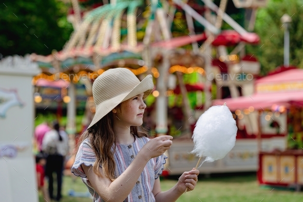 A girl in a hat and a dress eating cotton candy on a hot summer day at ...