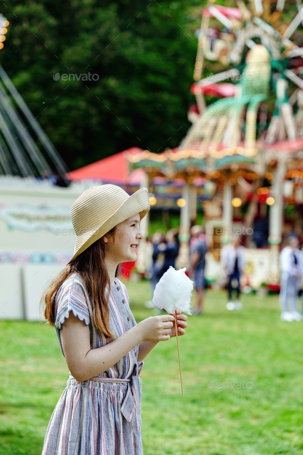A girl in a hat and a dress eating cotton candy on a hot summer day at ...