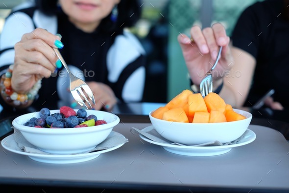 Two women sitting down eating fruits in the bowls by fork Stock Photo ...