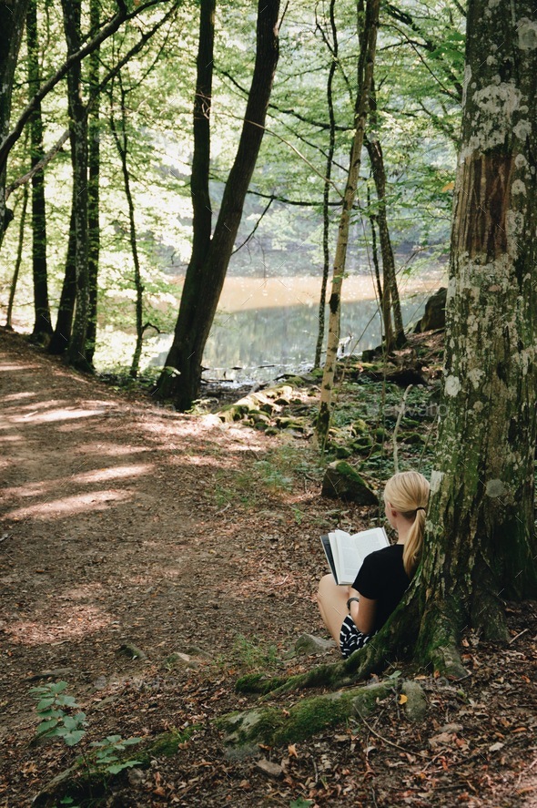 Photo of a girl reading in the forest, leaning her back against a tree ...