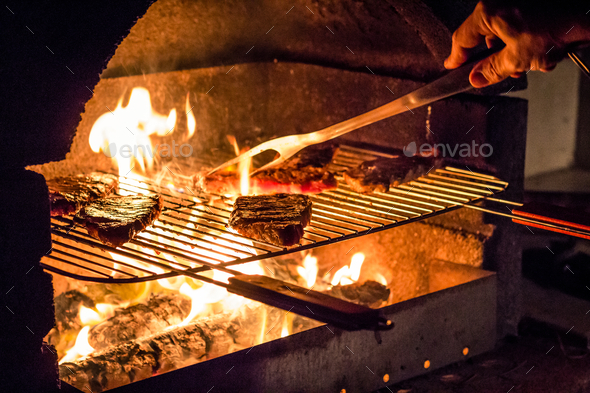 Hand holding a spindle in front of a grill with meat on it during a ...