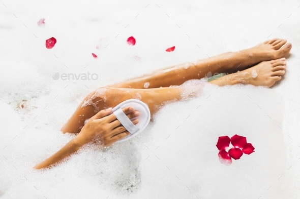 Woman washing legs in bath tub with foam bubbles and use natural sponge ...