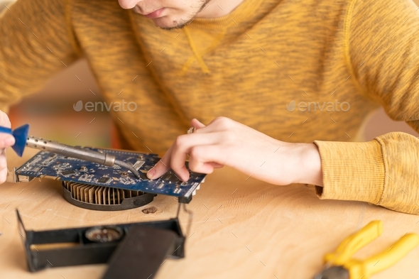 a young man solders a burnt-out microcircuit with a soldering iron ...
