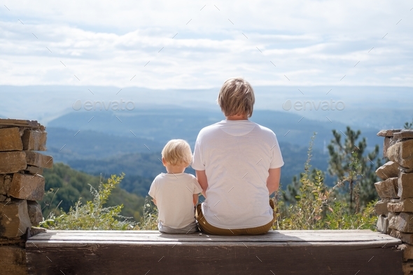 rear view father and son sitting together outdoors on the bench and ...