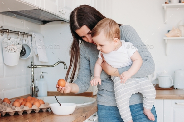 Young mother brunette woman with baby boy in arms cooking food in white ...