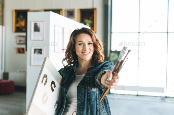 Charming young woman artist in denim jacket with paint brushes and ...