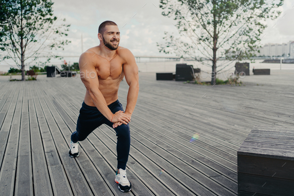 Horizontal shot of bearded motivated man stretches leg muscle before ...