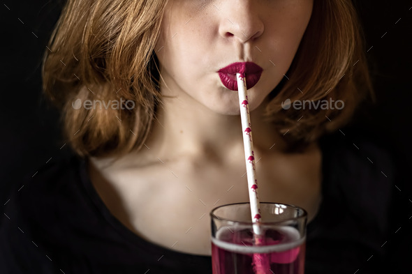 A young girl with bright pink lipstick drinks a cocktail from a glass ...
