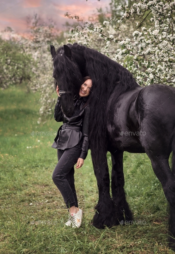 beautiful caucasian girl stands in blooming gardens with friesian horse ...