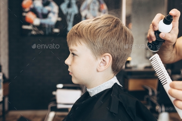 cute boy is sitting in a barbers chair and getting haircut and styling ...