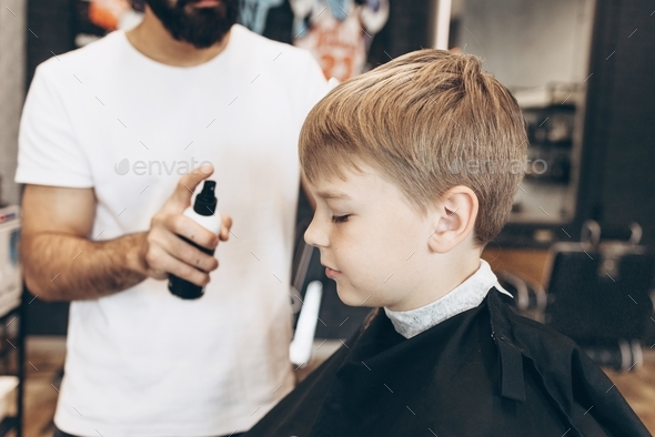 cute boy is sitting in a barbers chair and getting haircut and styling ...