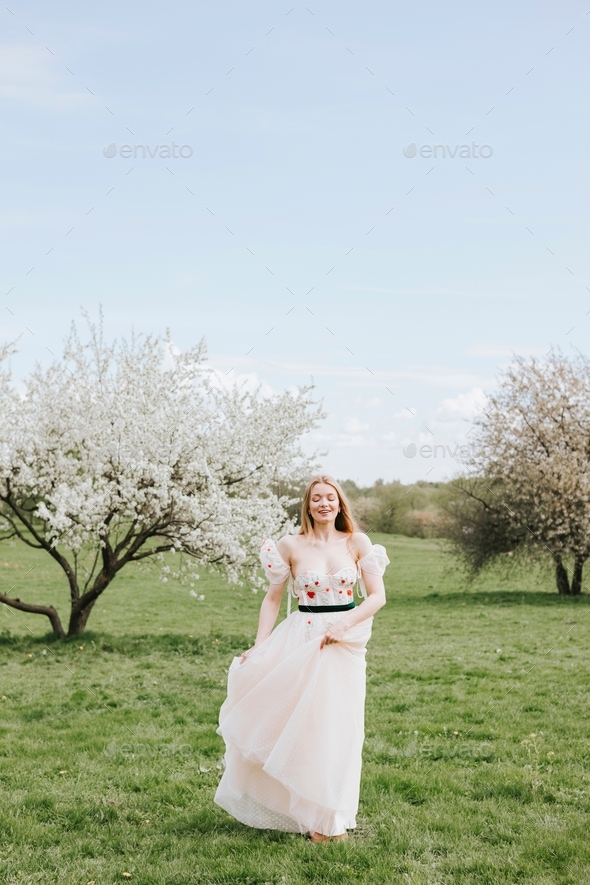 young beautiful blonde girl in long white dress poses near cherry ...