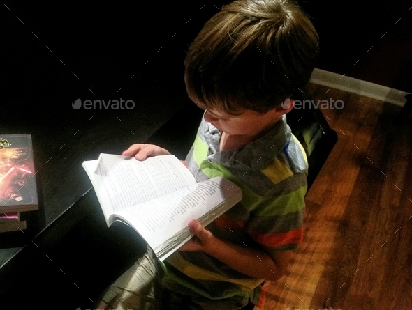 Little boy reading in low light because he supposed to be in bed ...