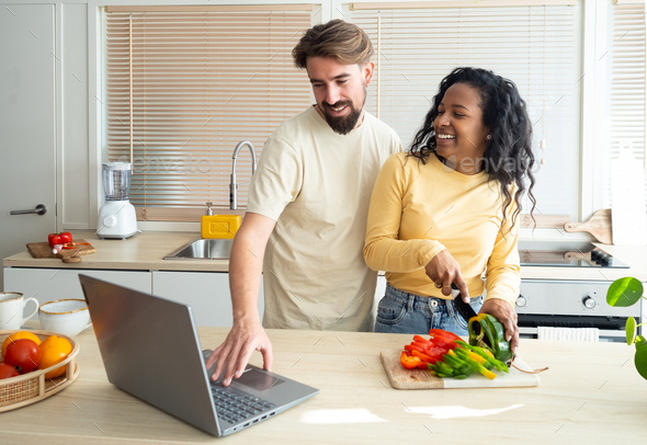 Happy multiethnic couple cooking together while watching cooking ...