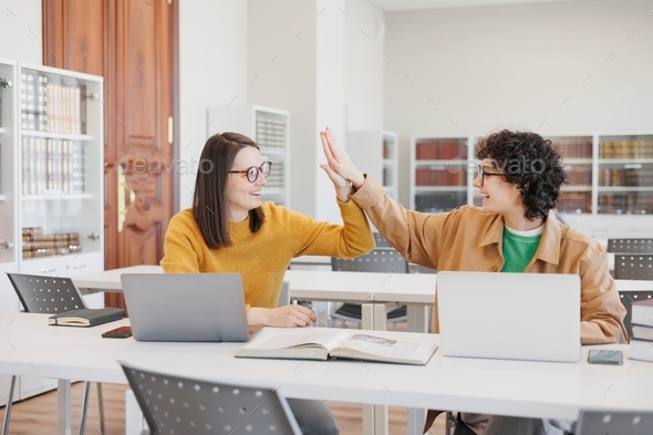 two women smile and give high fives working on a laptop, doing a ...