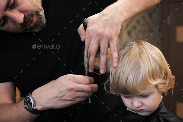 A little blonde boy is trimmed in the barbershop. Bright emotions on ...