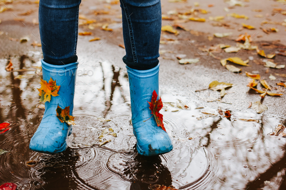 Teenager girl wearing blue rain boots jumping into a puddle on rainy autumn day. Closeup. N ...