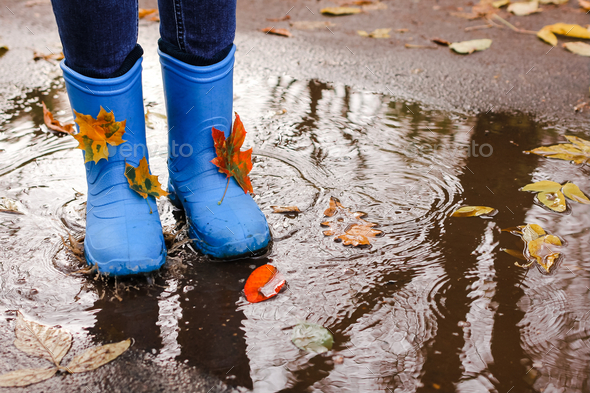 Teenager girl wearing blue rain boots jumping into a puddle on rainy autumn day. Closeup. N ...