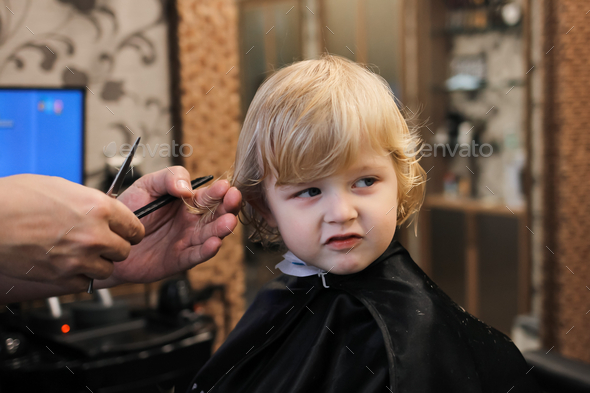 A little blonde boy is trimmed in the barbershop. Bright emotions on ...