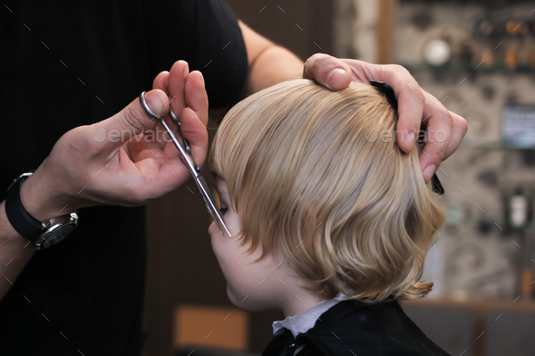 A little blonde boy is trimmed in the barbershop. Bright emotions on ...