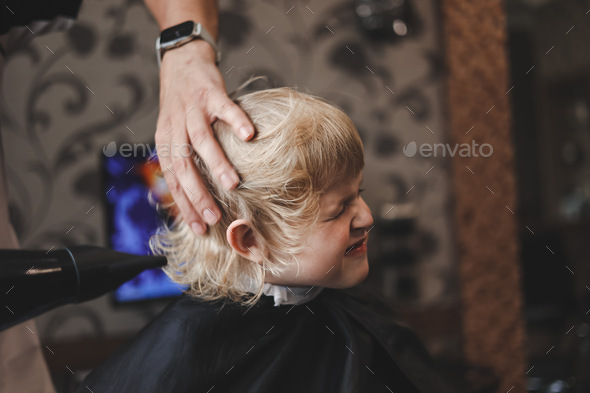 A little blonde boy is trimmed in the barbershop. Bright emotions on ...