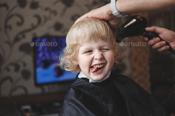A little blonde boy is trimmed in the barbershop. Bright emotions on ...