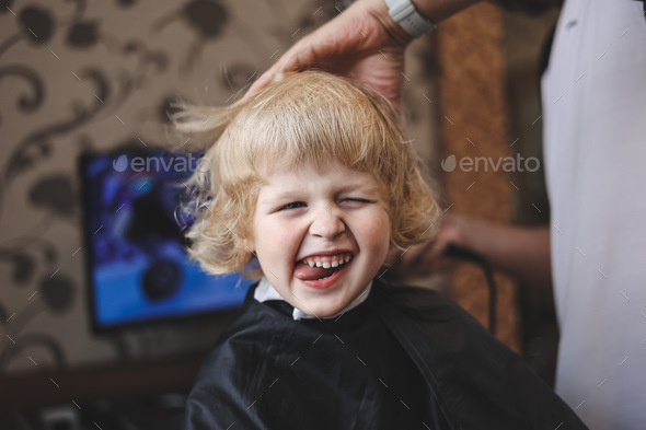A little blonde boy is trimmed in the barbershop. Bright emotions on ...