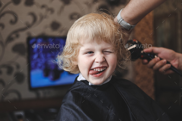 A little blonde boy is trimmed in the barbershop. Bright emotions on ...