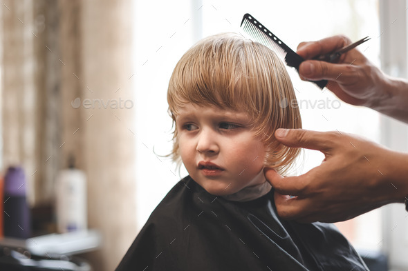A little blonde boy is trimmed in the barbershop. Bright emotions on ...