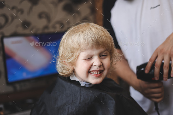 A little blonde boy is trimmed in the barbershop. Bright emotions on ...