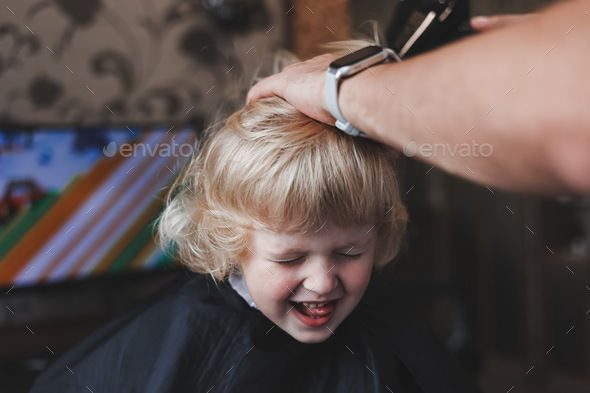 A little blonde boy is trimmed in the barbershop. Bright emotions on ...