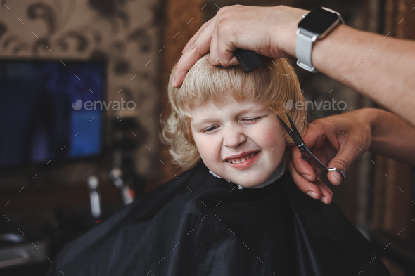 A little blonde boy is trimmed in the barbershop. Bright emotions on ...