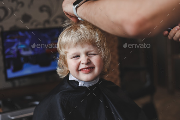 A little blonde boy is trimmed in the barbershop. Bright emotions on ...