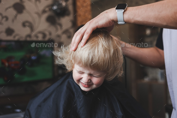 A little blonde boy is trimmed in the barbershop. Bright emotions on ...