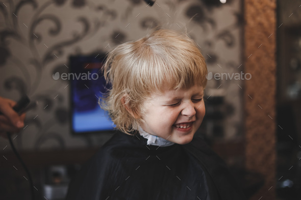 A little blonde boy is trimmed in the barbershop. Bright emotions on ...