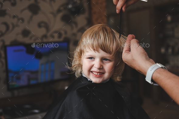 A little blonde boy is trimmed in the barbershop. Bright emotions on ...