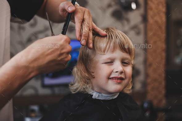A little blonde boy is trimmed in the barbershop. Bright emotions on ...