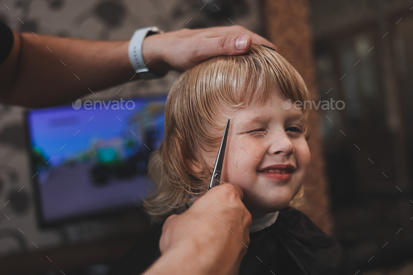 A little blonde boy is trimmed in the barbershop. Bright emotions on ...