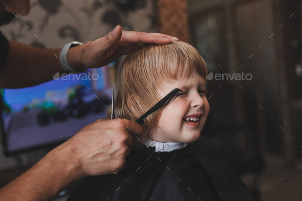 A little blonde boy is trimmed in the barbershop. Bright emotions on ...