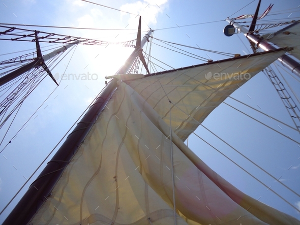 Looking up at cloth sails at the sun & blue skies on a tall mast ...